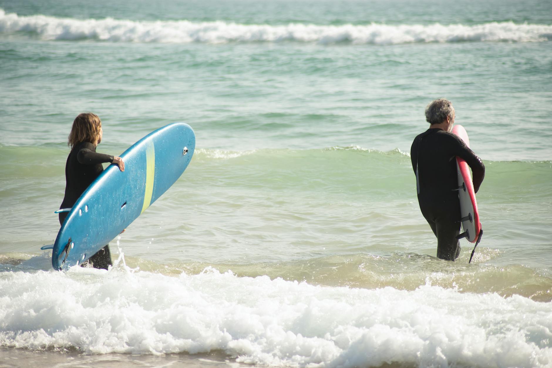 Two surfers with boards head into ocean waves on a bright day