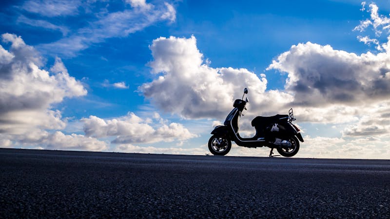 Silhouette of a Vespa scooter on an open road under bright blue sky