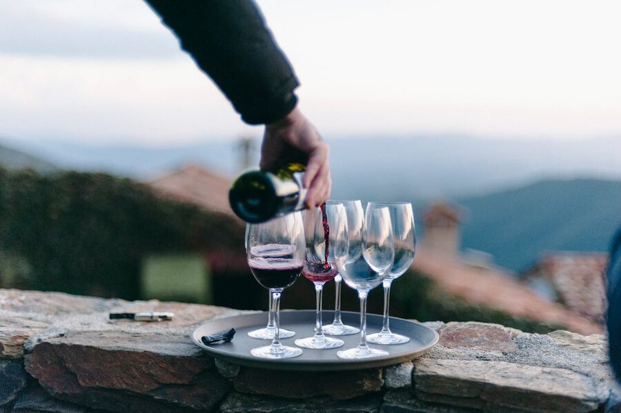Hand pouring red wine into glasses on a tray in an outdoor setting with hills in the background
