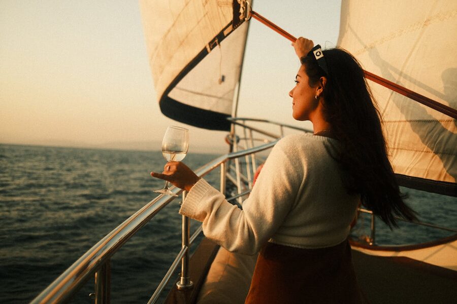 Woman stands on sailboat holding wine glass enjoying sunset view