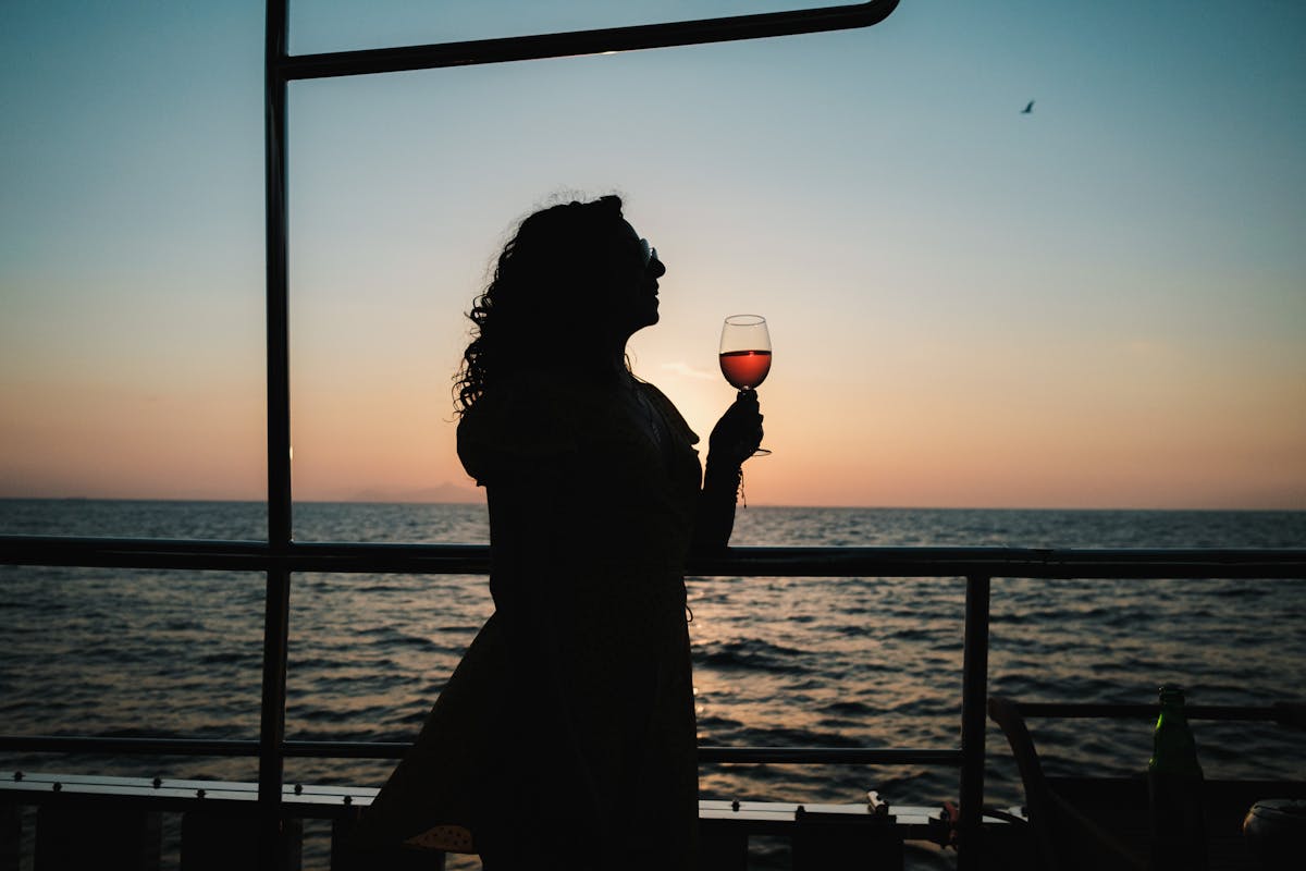 A woman silhouetted against a sunset over the ocean enjoying a glass of wine on a boat